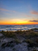 Uma praia com um pôr do sol sobre o oceano, dunas de areia e vegetação rala.