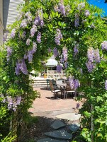 Um jardim com pérgola coberta de flores roxas, um alpendre com móveis de exterior e uma cerca branca.