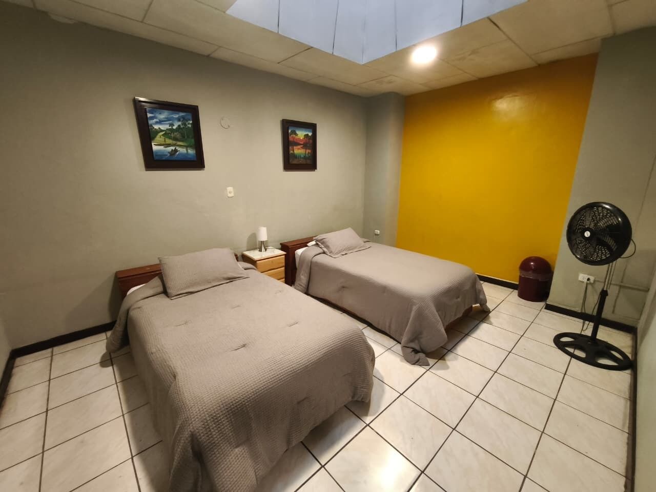 Quarto com duas camas, mesa de cabeceira, ventilador e duas fotos emolduradas na parede.