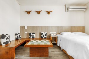 Quarto de hotel com cabeceira de madeira, cama com roupa de cama branca, um banco de madeira com um jogo de tabuleiro xadrez e um ar condicionado fixado na parede.