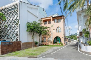 Rua estreita ladeada por edifícios residenciais, incluindo uma casa com uma grande escultura de girafa e um edifício branco com fachada treliçada.