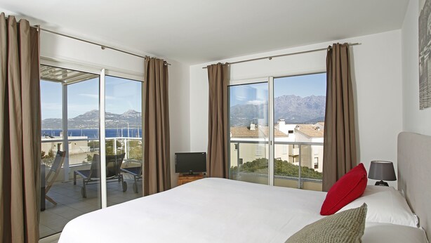 Quarto de hotel com uma cama grande, TV e varanda com vista para o mar e montanhas.