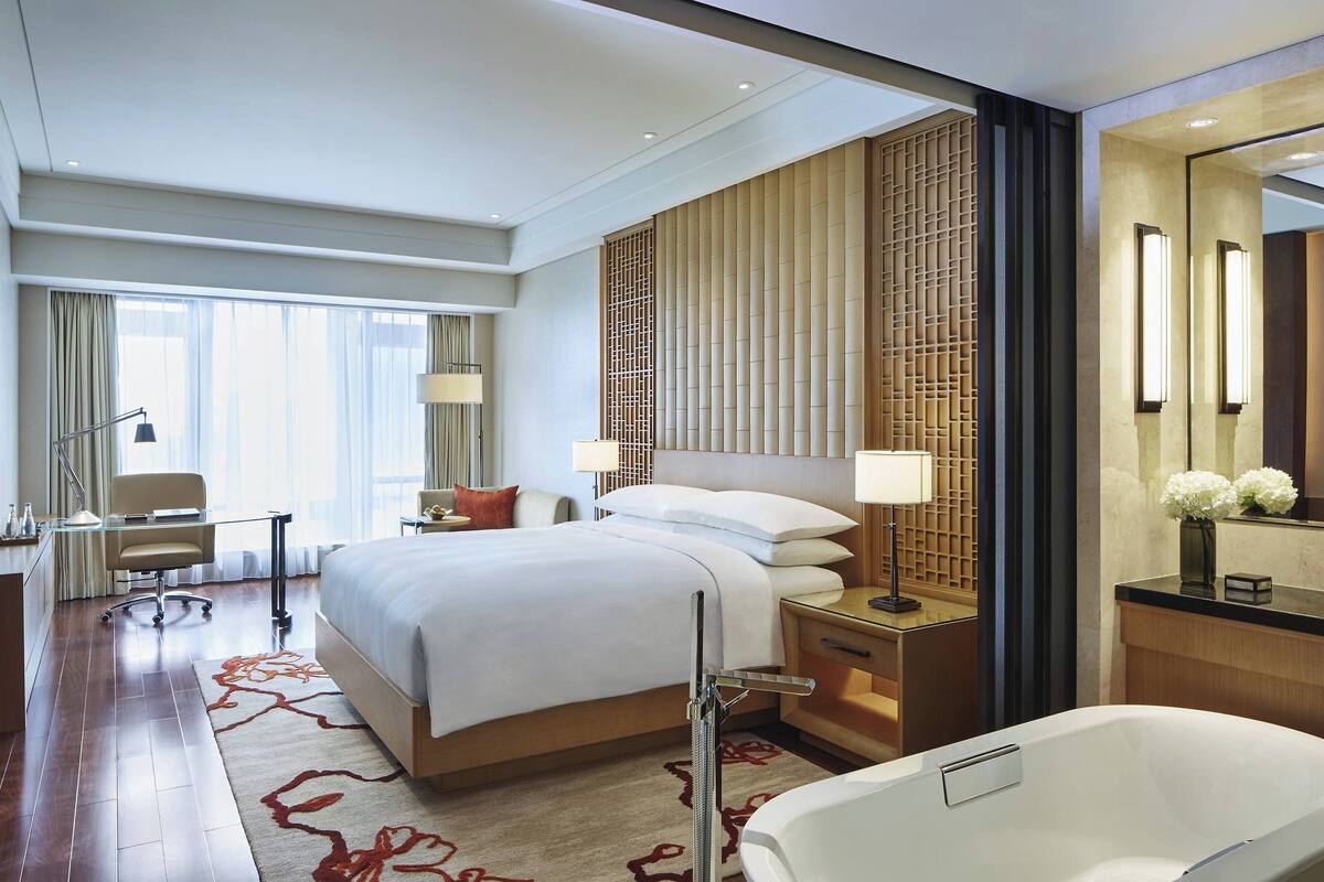 Quarto de hotel moderno com uma cama grande, um espaço de estar com uma poltrona e escrivaninha, banheiro com banheira e uma janela ampla com cortinas.