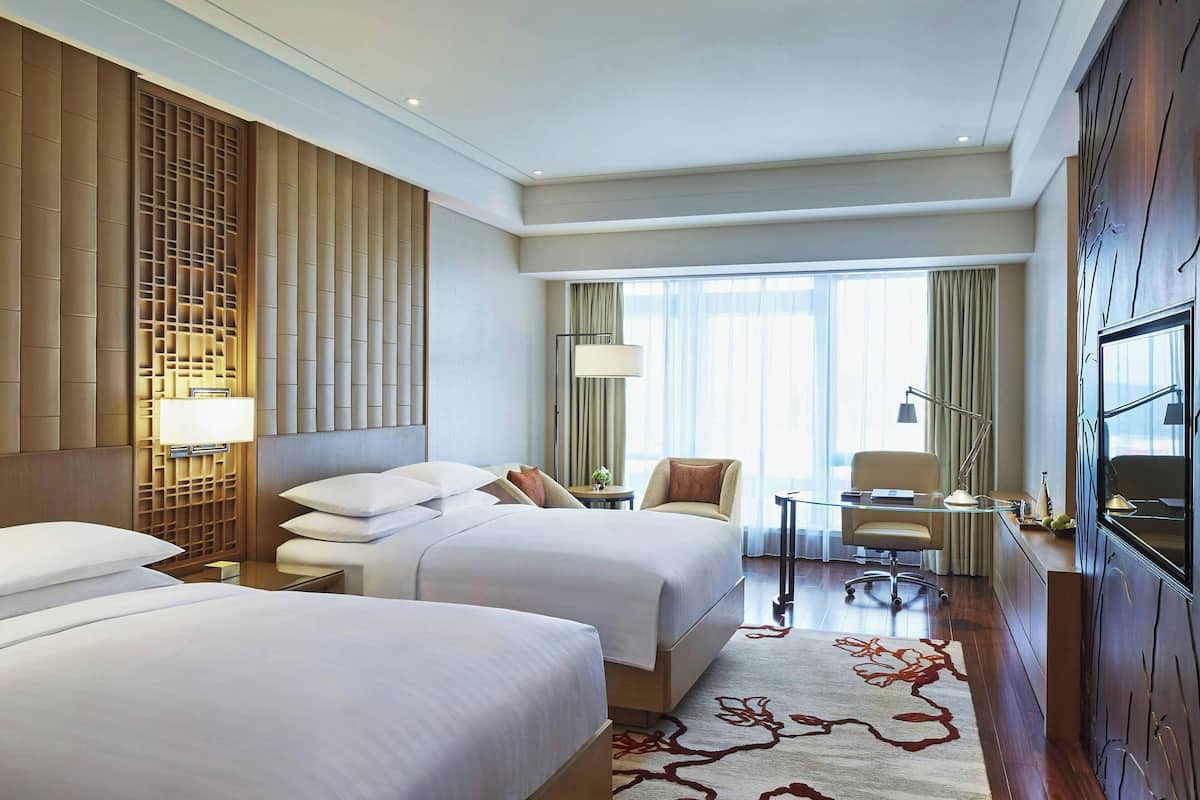 Quarto de hotel com duas camas, uma escrivaninha, uma cadeira, televisão e uma janela grande com cortinas.