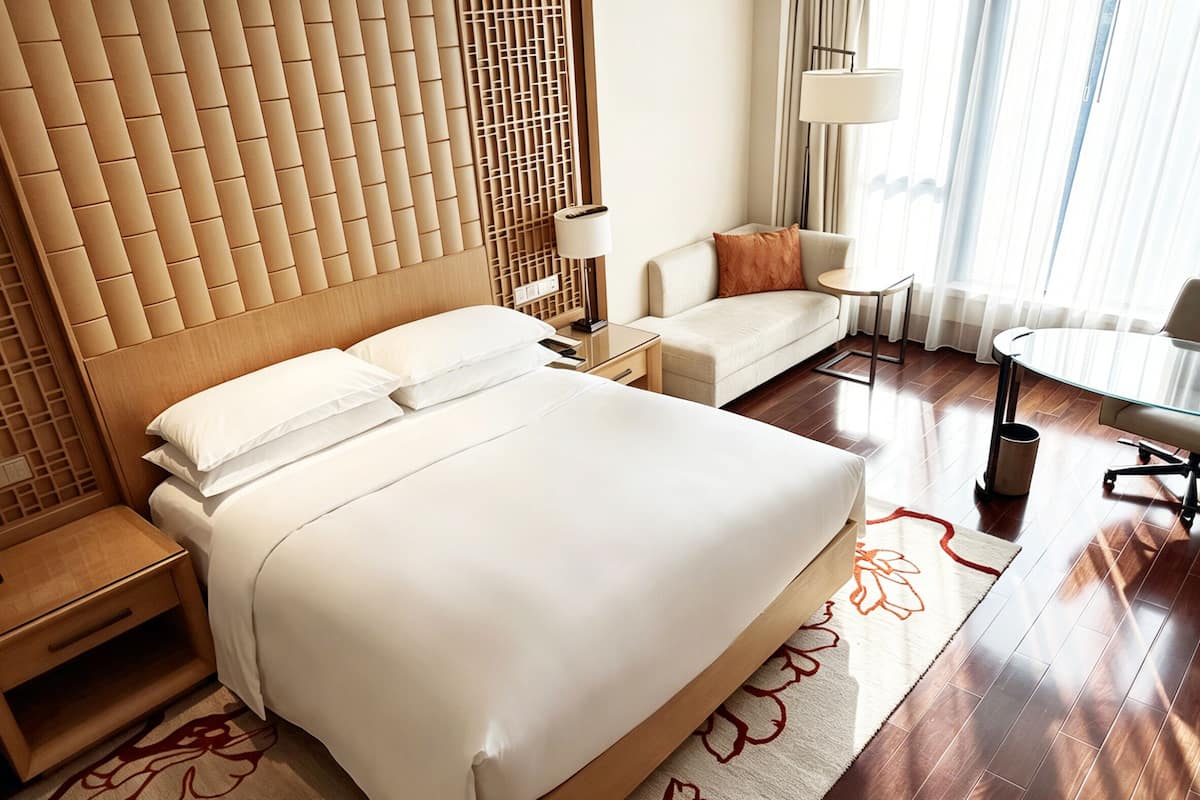 Quarto de hotel com uma cama grande, mesa de cabeceira, sofá, pequena mesa, abajur e vista para o exterior.