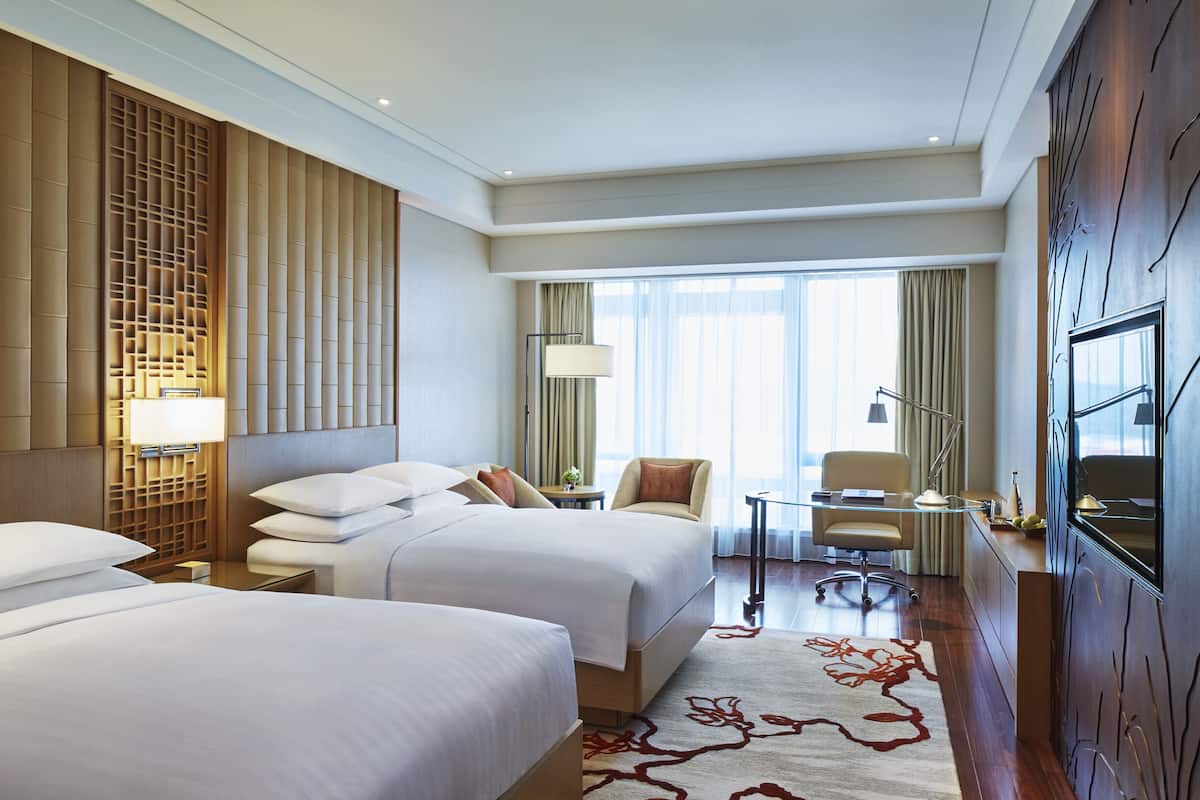 Quarto de hotel com duas camas, uma mesa, uma cadeira, uma televisão e uma janela grande.