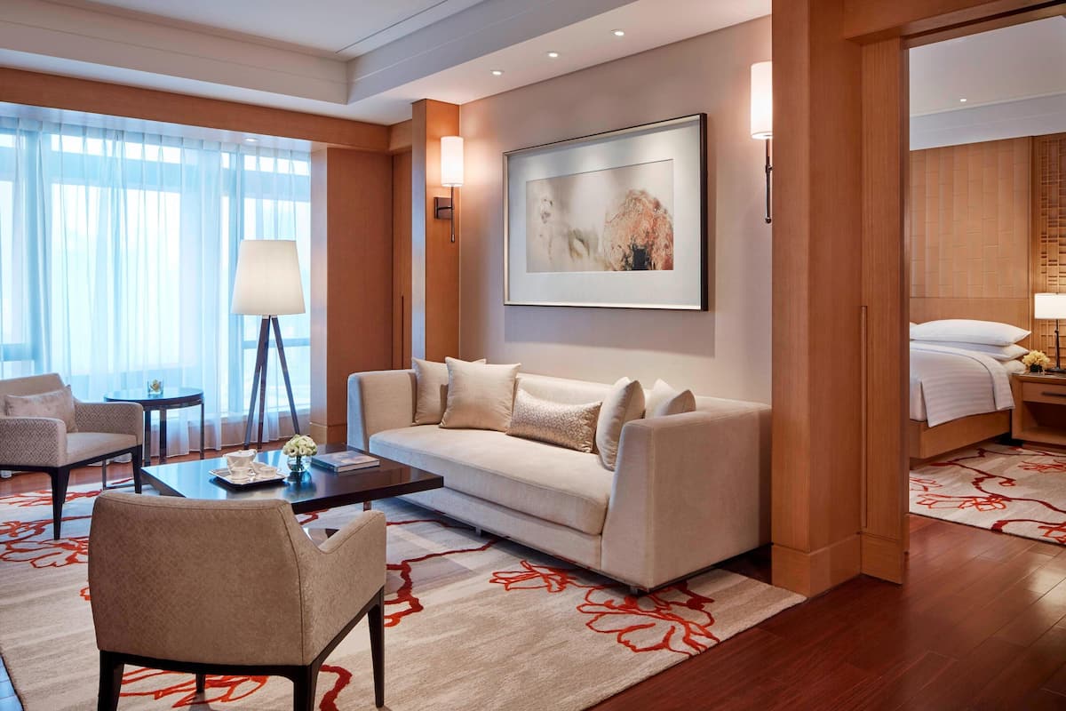 Quarto de hotel moderno com sofá, poltrona e mesa de centro. Há um quadro na parede e uma cama com mesa de cabeceira.