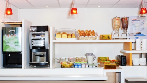 Uma área de recepção de hotel com máquina de café, frigorífico, cesta de frutas e um buffet com pão e pastelaria.