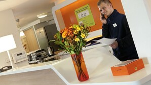 Balcão de recepção de hotel com um vaso de flores, uma etiqueta de identificação e um aviso sobre tarifas de quartos.