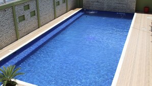 Uma piscina retangular com água azul cristalina, rodeada por um deck ladrilhado e uma parede de pedra com detalhes verdes.
