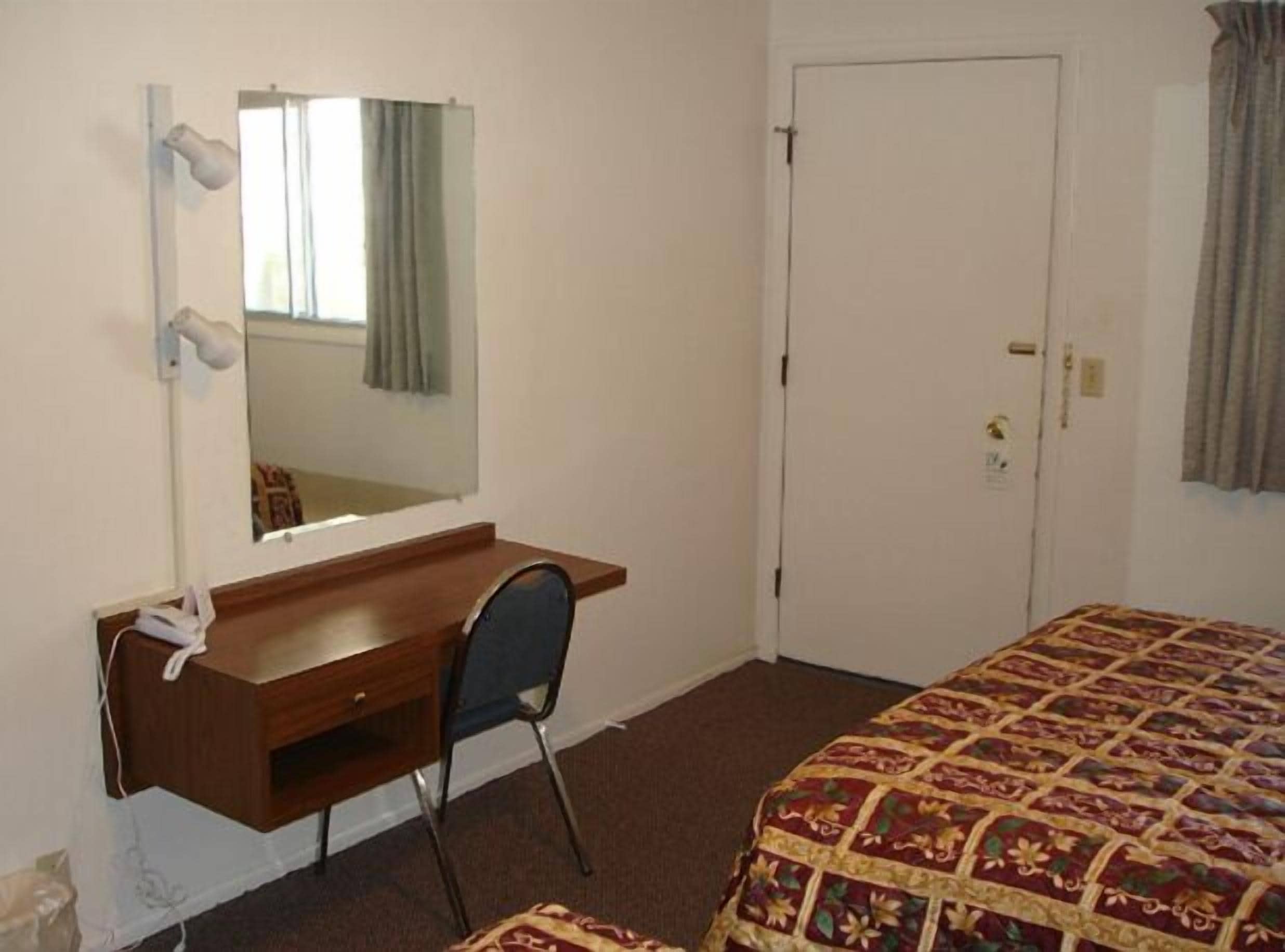 Quarto de hotel com escrivaninha de madeira, cadeira, cama com colcha estampada, espelho e porta.