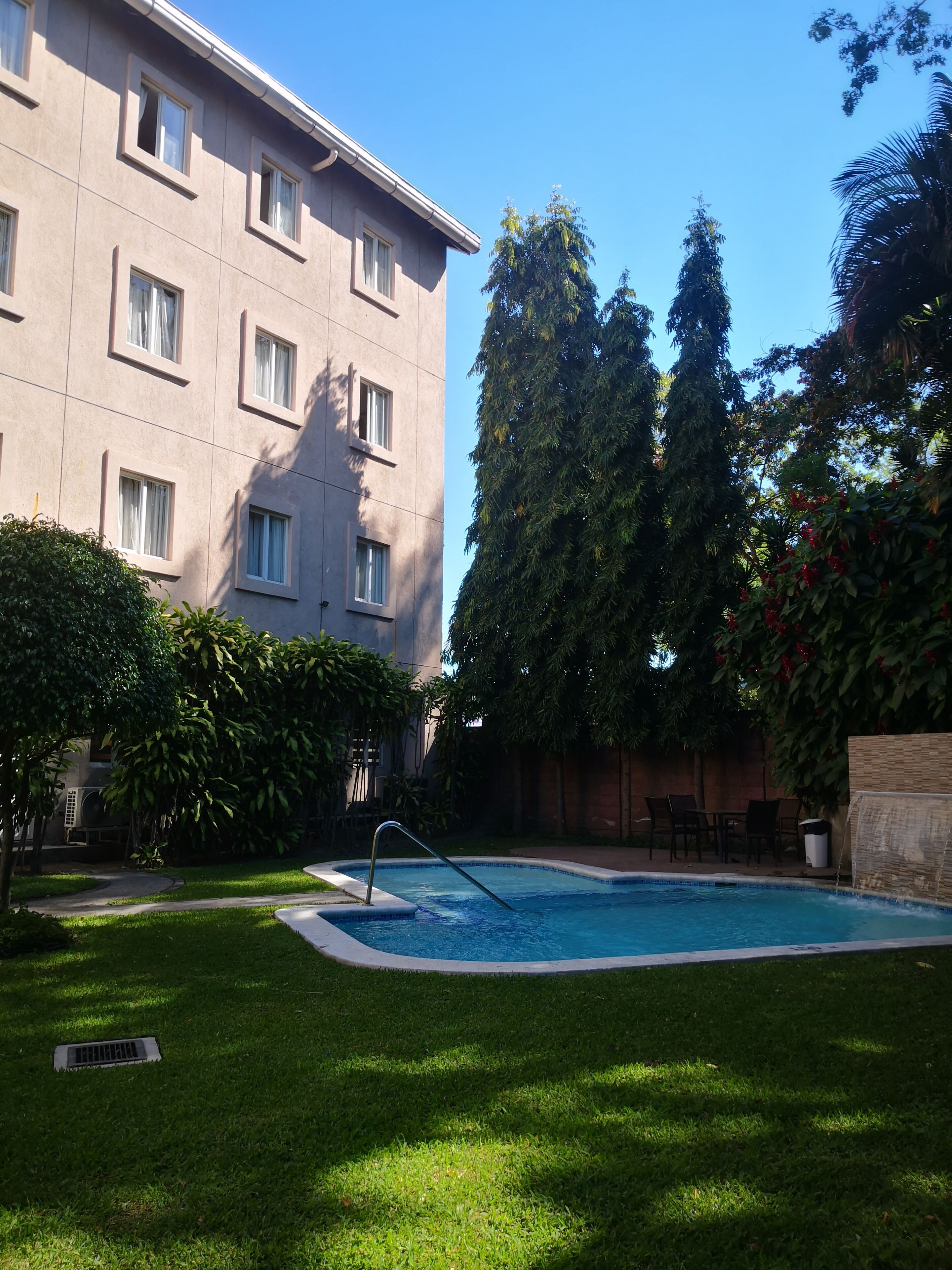Uma piscina rodeada por um espaço gramado, com um edifício ao fundo.