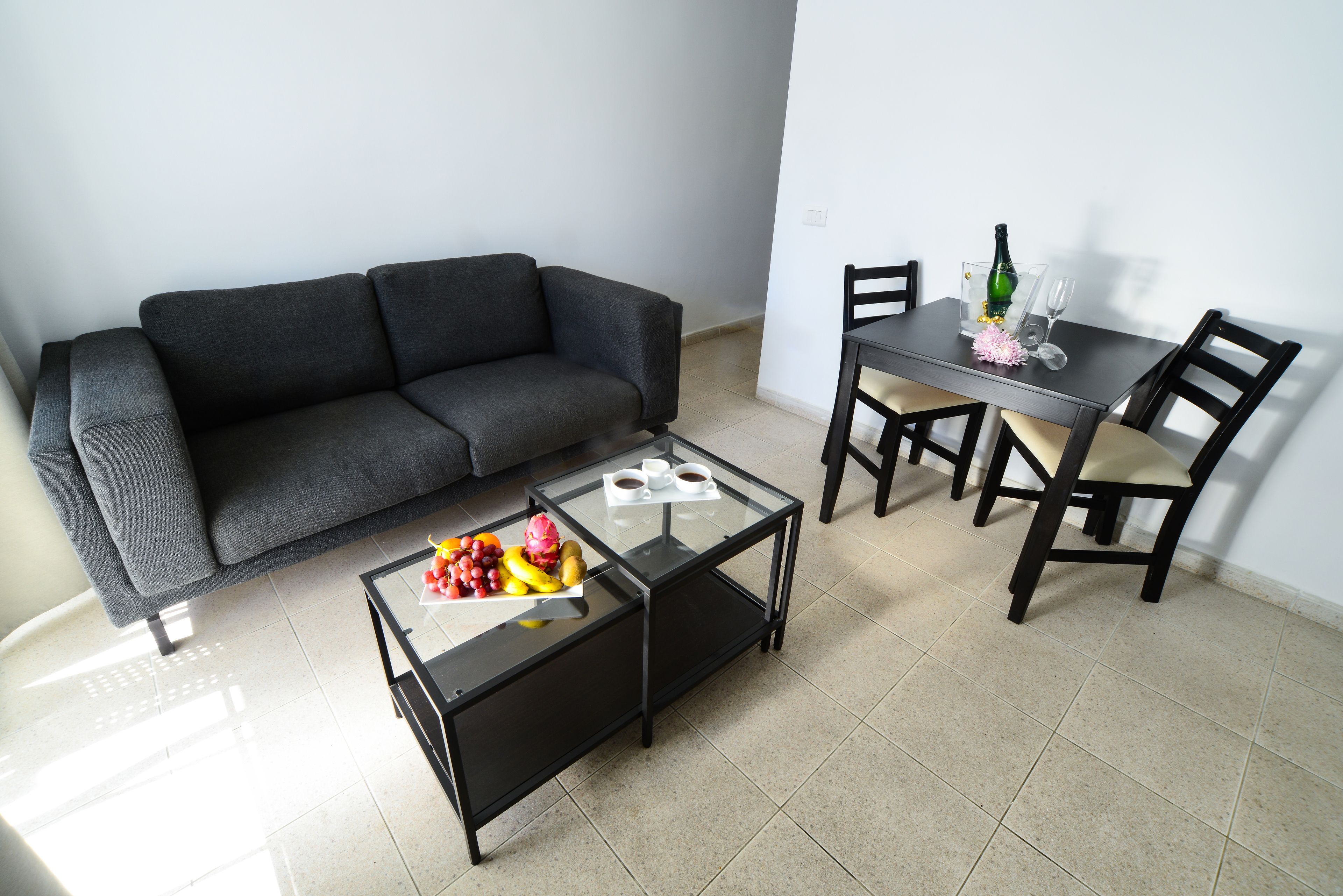 Uma sala de estar com sofá, mesa de centro com frutas, mesa de vidro com garrafa e copo, e mesa de jantar com cadeiras.