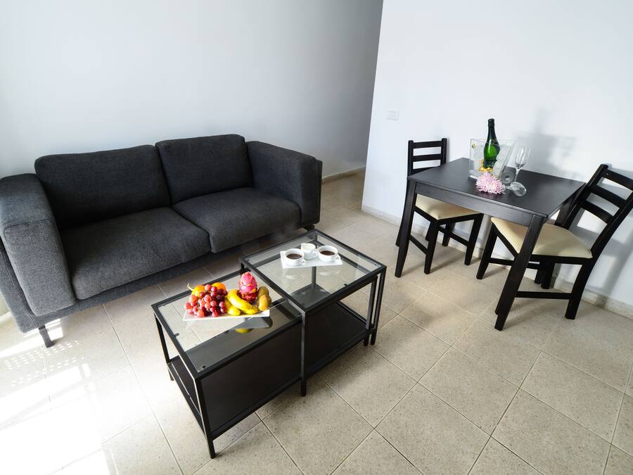 Uma sala de estar com sofá, mesa de centro com frutas, mesa de vidro com garrafa e copo, e mesa de jantar com cadeiras.
