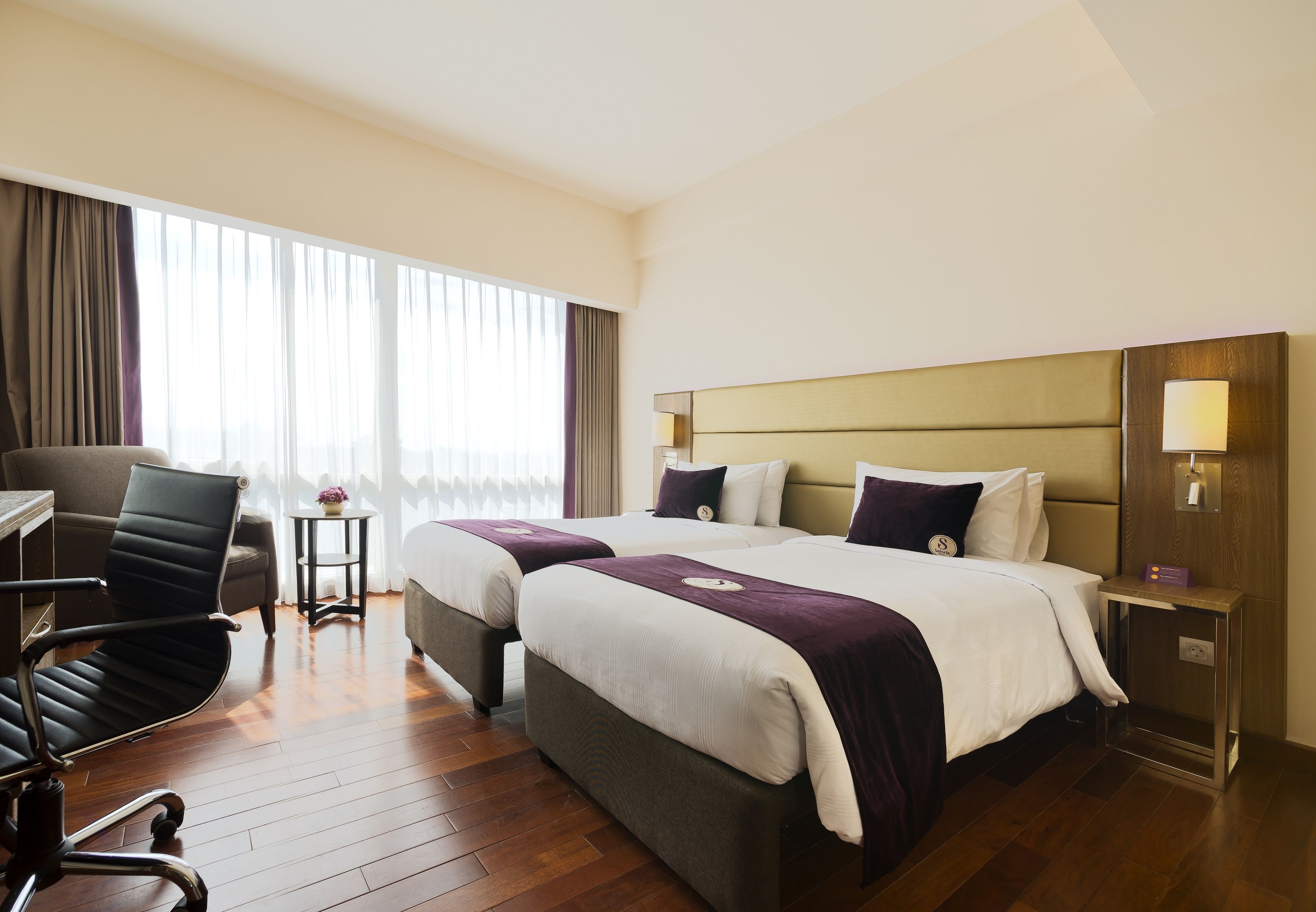 Quarto de hotel com duas camas, uma escrivaninha, uma cadeira e uma mesa de cabeceira com abajur.