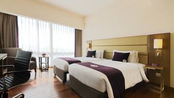Quarto de hotel com duas camas, uma escrivaninha, uma cadeira e uma mesa de cabeceira com abajur.