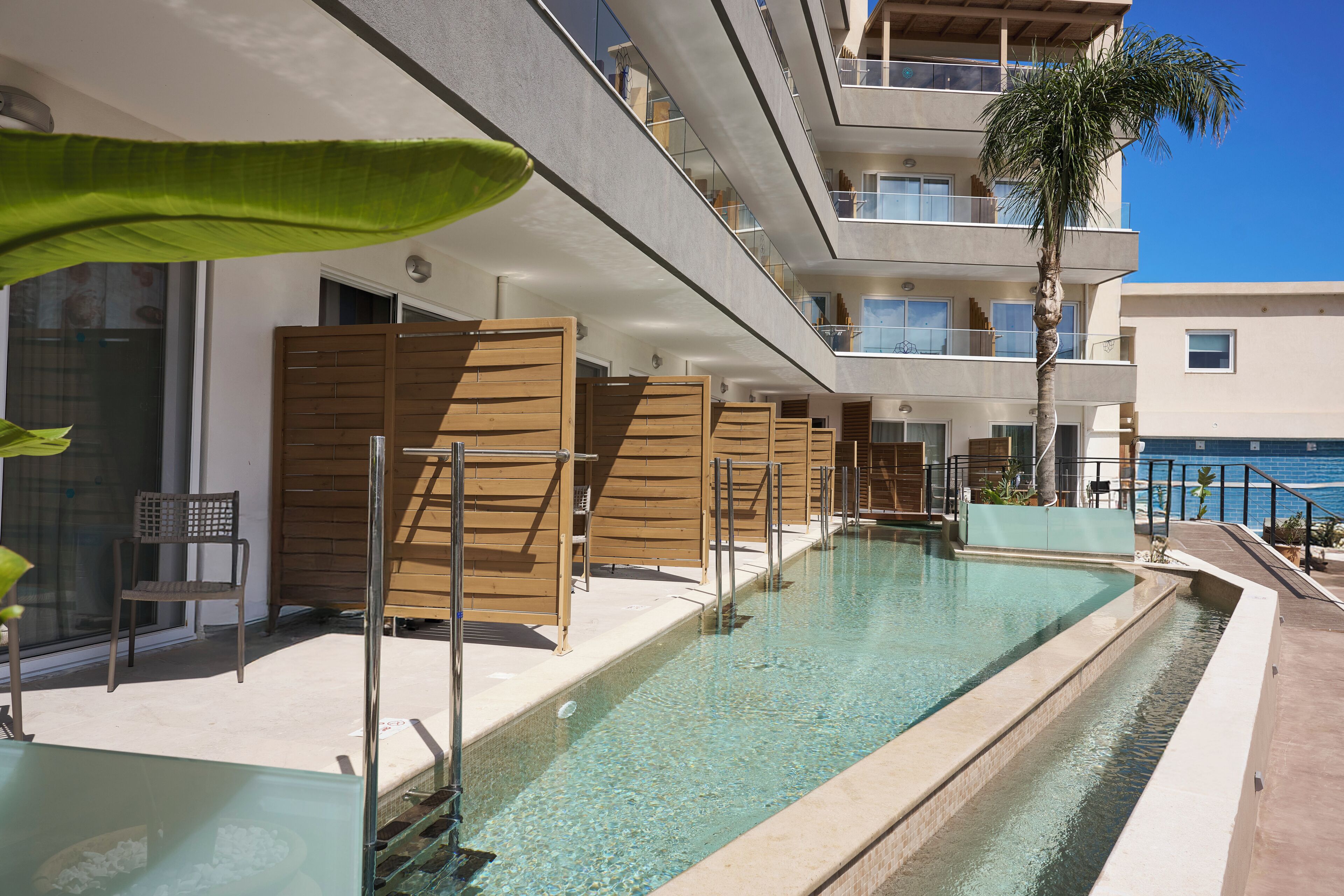 Piscina com espreguiçadeiras de madeira, uma palmeira e um prédio com várias andares e sacadas.