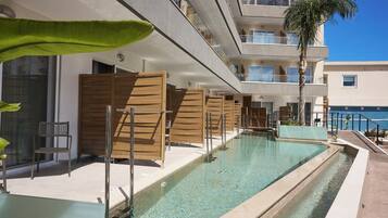 Piscina com espreguiçadeiras de madeira, uma palmeira e um prédio com várias andares e sacadas.
