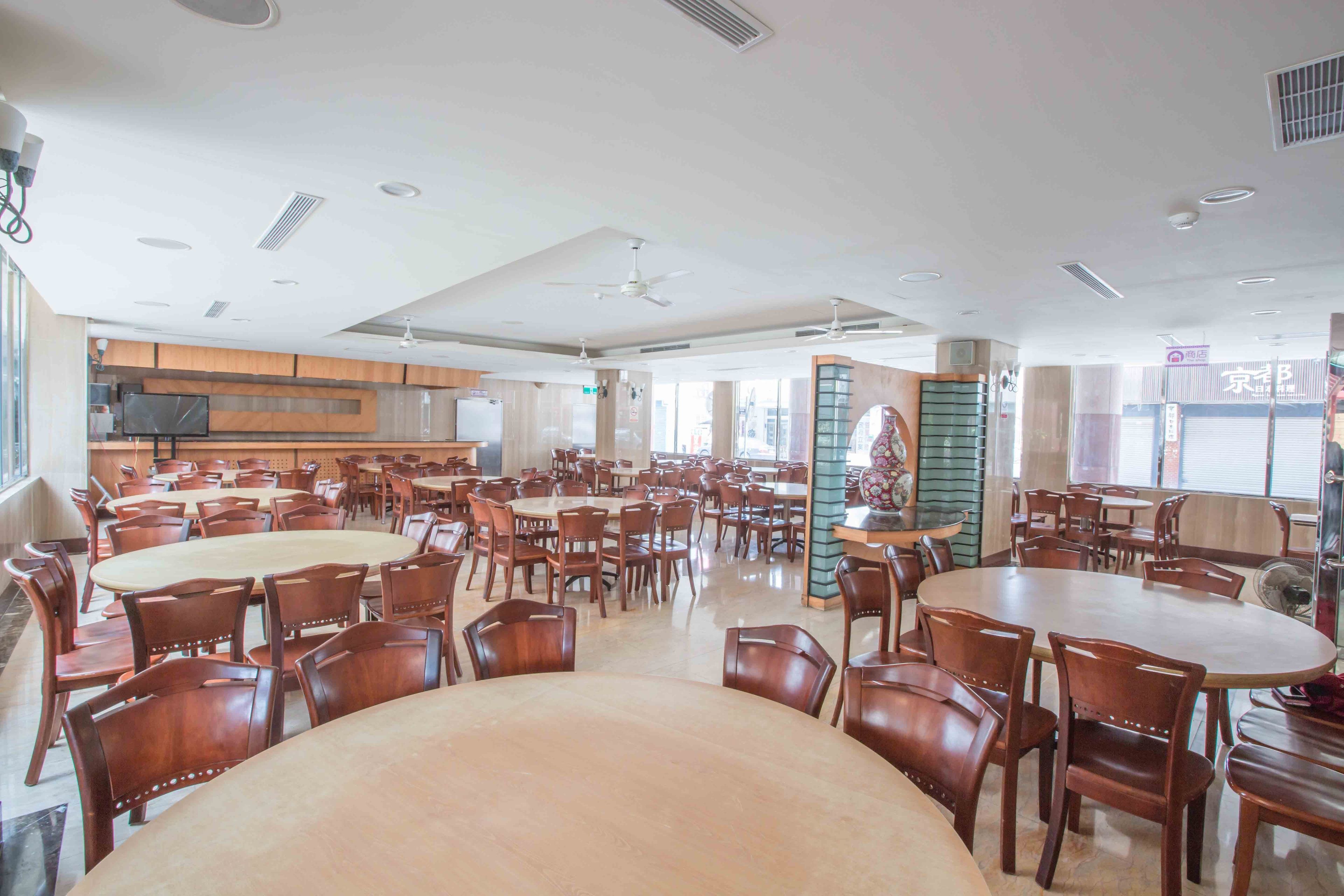Uma sala de jantar ampla com várias mesas redondas e cadeiras, ventiladores de teto e uma grande janela que proporciona iluminação natural.