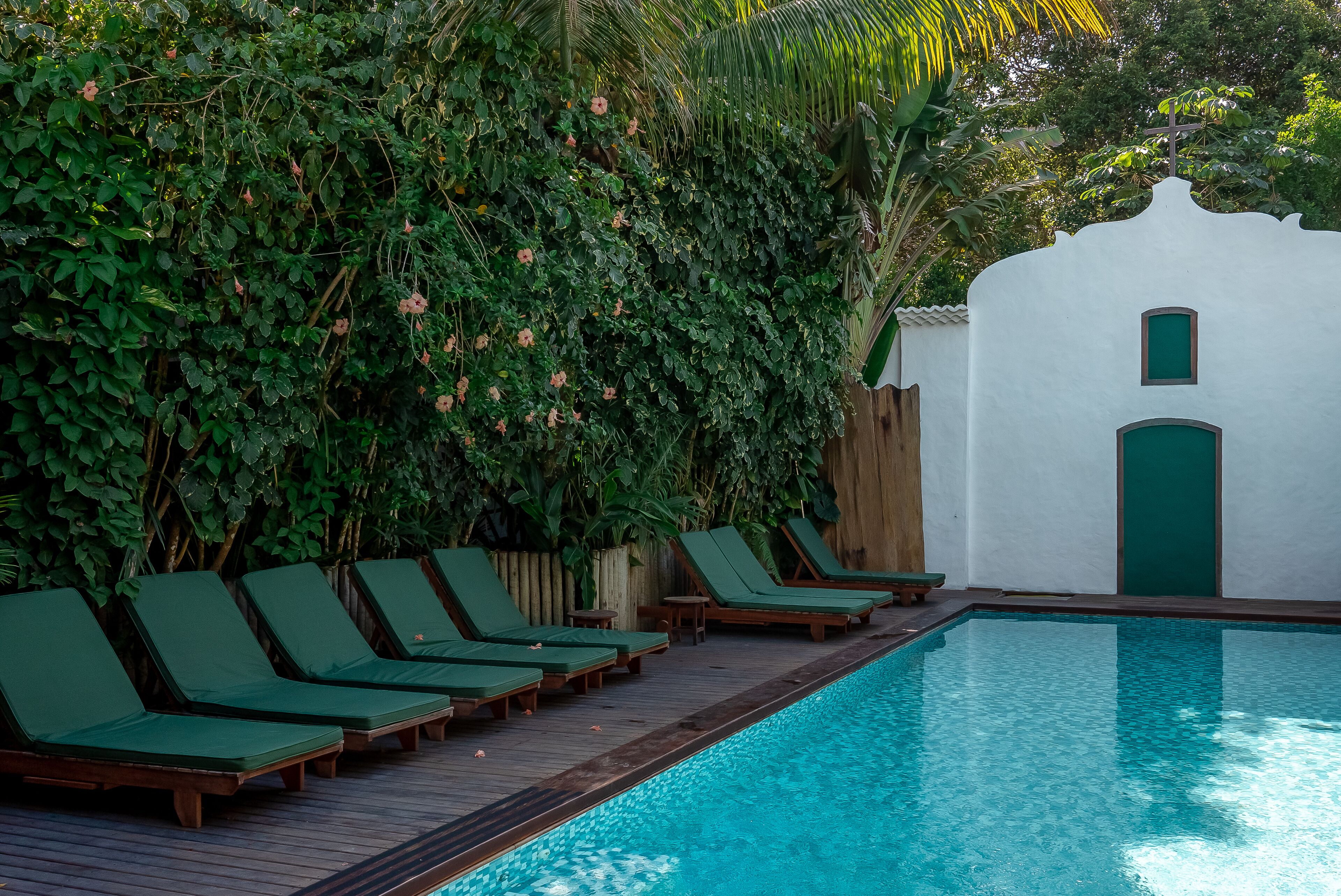 Área de piscina com espreguiçadeiras de madeira e uma parede branca com uma porta verde.