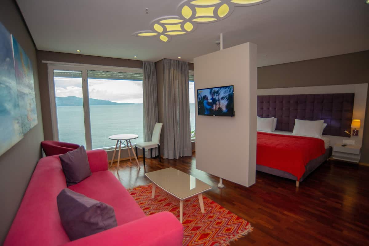 Quarto de hotel moderno com uma cama grande, um sofá vermelho, uma mesa pequena, uma TV e vista para o mar através de grandes janelas.