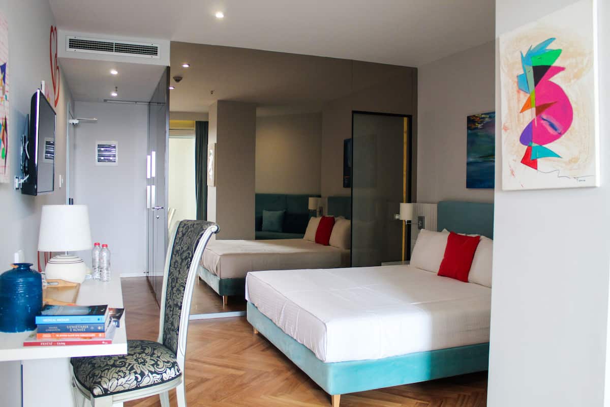 Quarto de hotel com duas camas, uma escrivaninha com cadeira, televisão e uma janela grande.