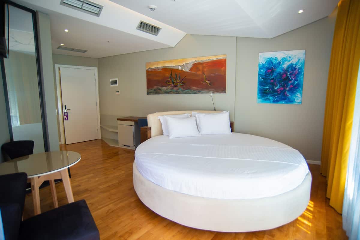 Quarto de hotel moderno com uma cama redonda grande, uma pequena escrivaninha, uma cadeira e duas pinturas na parede.