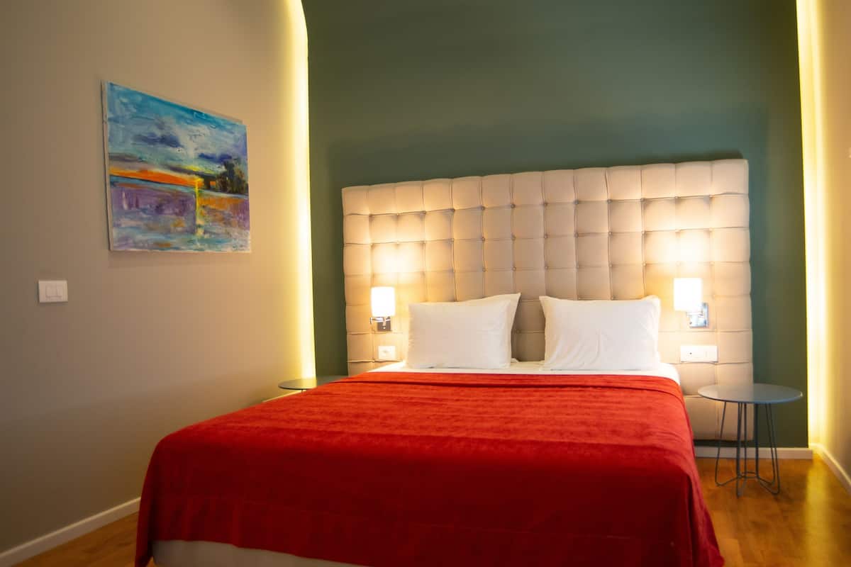 Quarto de hotel com uma cama grande, cabeceira com iluminação, uma mesinha e um quadro na parede.