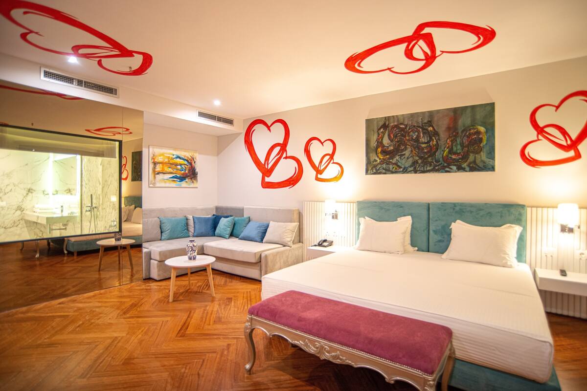 Quarto de hotel com uma cama grande, um sofá, uma mesinha e um banheiro visível por meio de um painel de vidro.