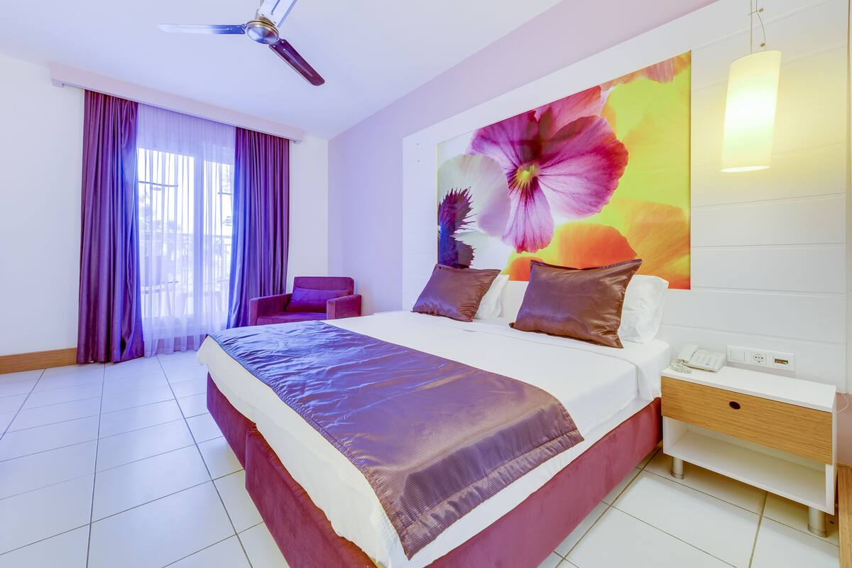 Quarto de hotel com uma cama grande, mesa de cabeceira, cadeira, ventoinha no teto e um mural floral na parede.