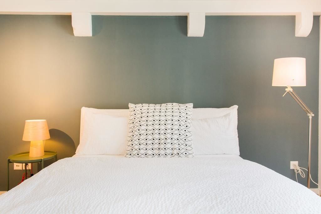 Cama bem arrumada com edredom branco, almofada estampada e abajur sobre um criado-mudo verde.