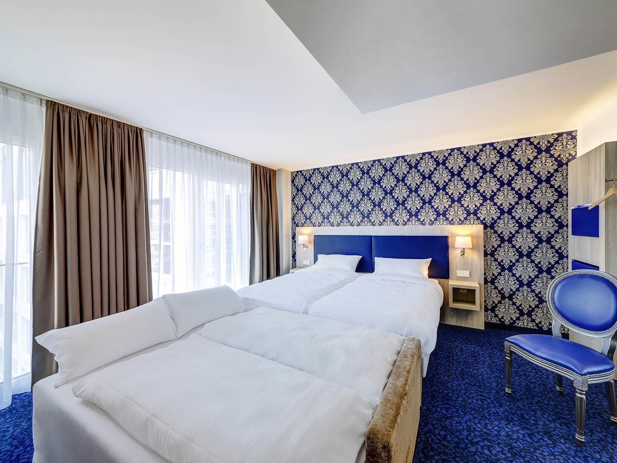 Quarto de hotel com uma cama grande, uma cadeira, uma mesa de cabeceira e um papel de parede estampado.