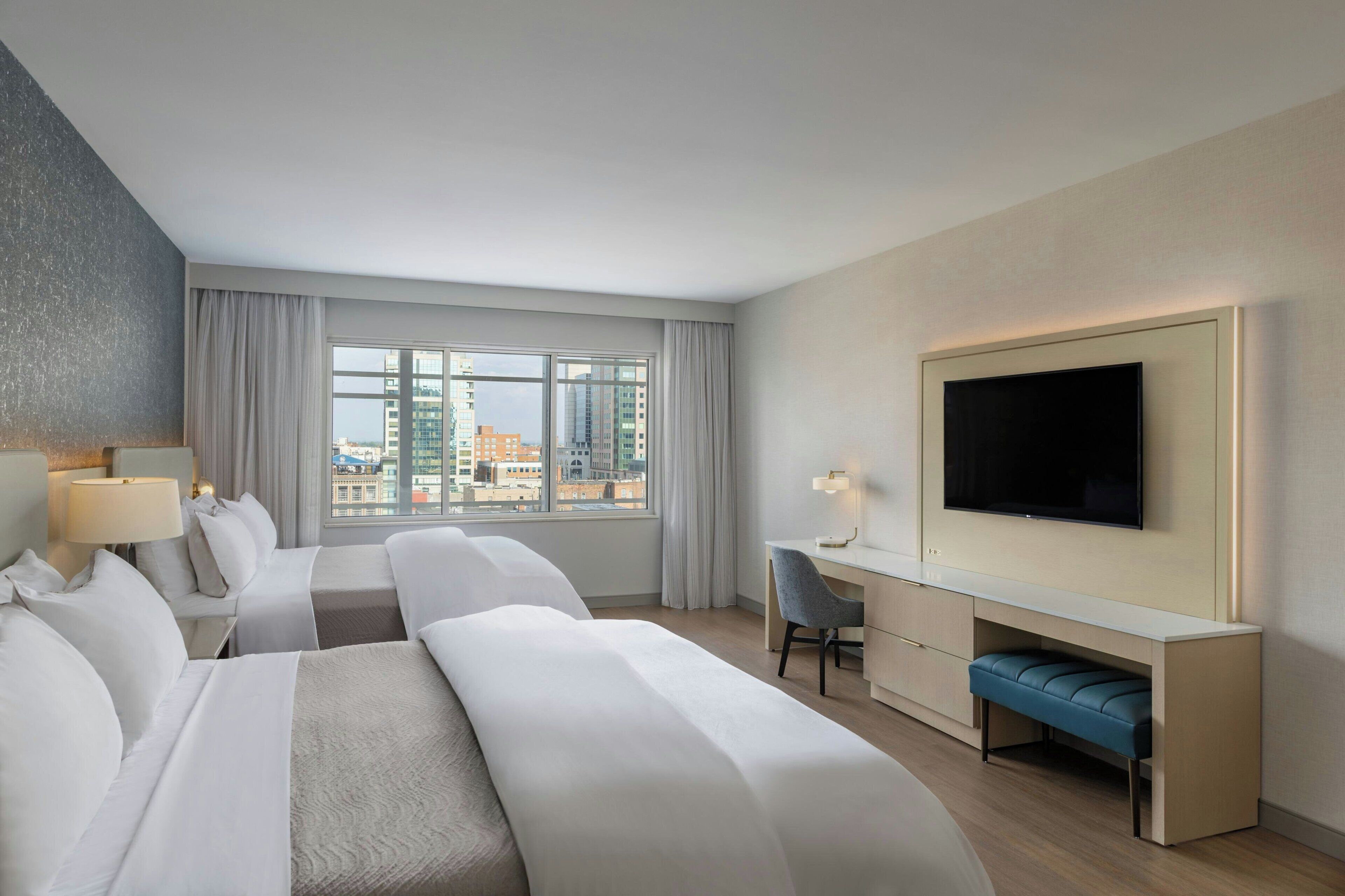 Quarto de hotel com duas camas, uma escrivaninha, uma cadeira, um banco, uma televisão e uma janela grande com vista para a cidade.