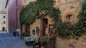Uma rua estreita de paralelepípedos com um edifício rústico, apresentando um arco de tijolos e paredes cobertas de hera. Há uma cadeira de madeira e uma mesa com toalha xadrez do lado de fora.