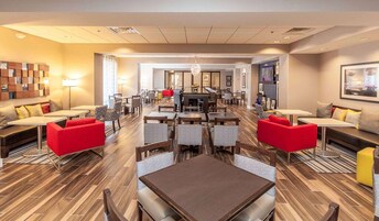 Um amplo saguão de hotel com área de estar central, várias mesas e uma lareira.
