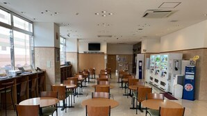 Uma cafeteria ampla com mesas e cadeiras redondas, máquinas de venda automática e uma grande janela que oferece luz natural.
