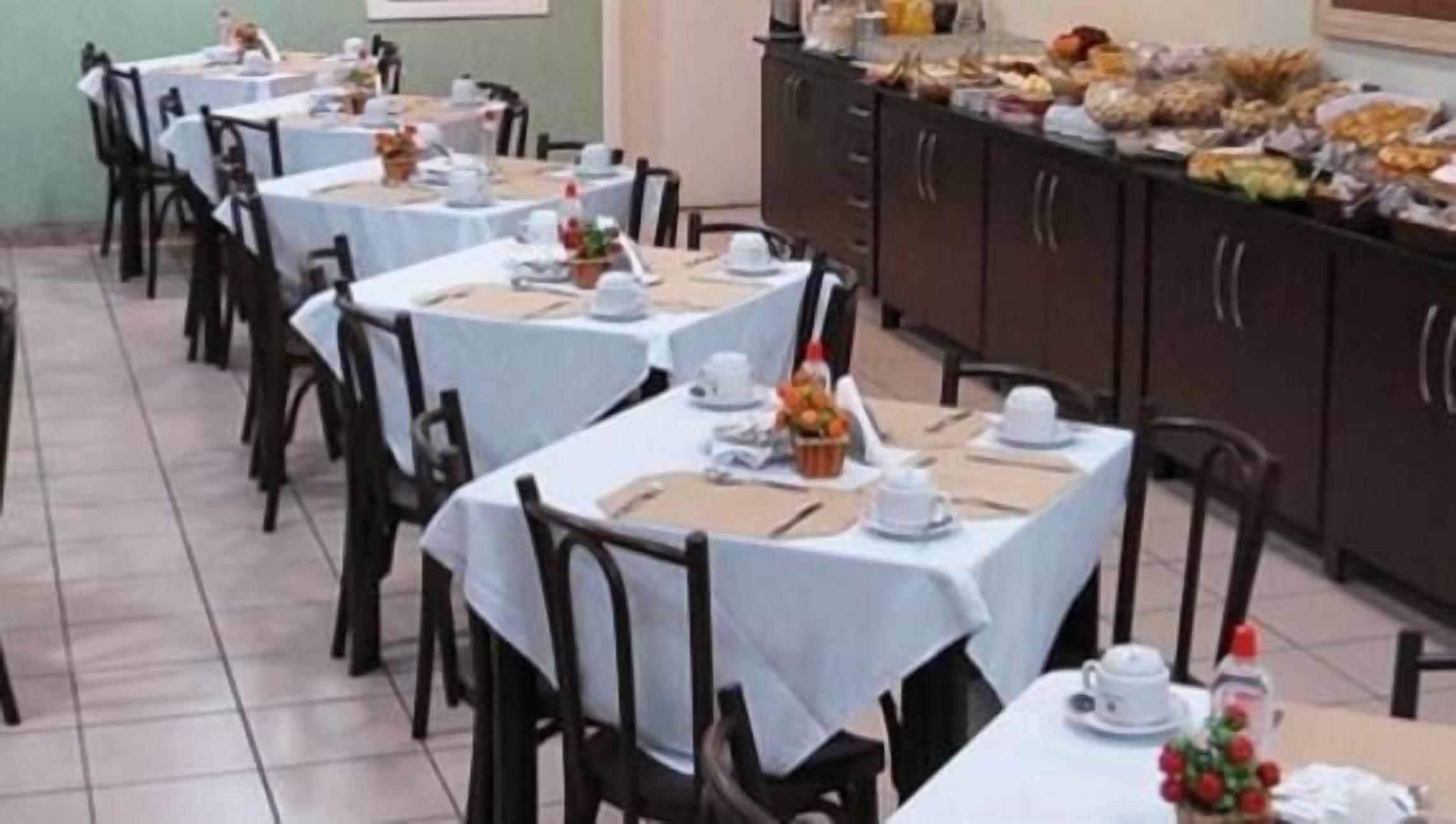 Uma área de refeições com mesas preparadas para o café da manhã, incluindo pratos, xícaras e diversos alimentos em um balcão de buffet.