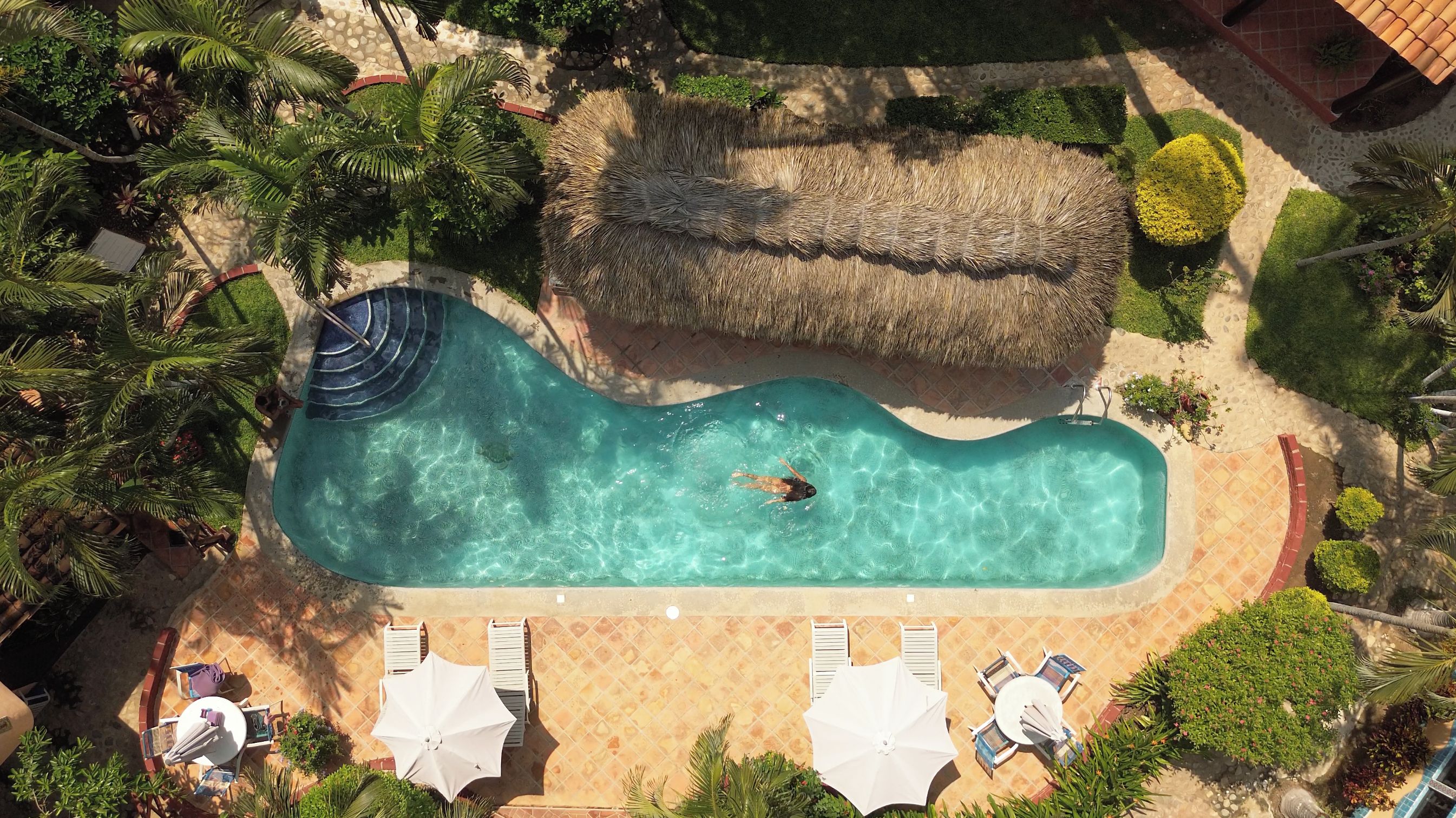 Vista aérea de uma piscina com alguém nadando, cercada por cadeiras de praia e guarda-sóis, e uma estrutura com teto de palha.