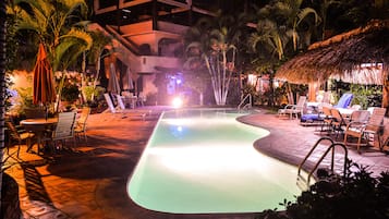 Piscina com espreguiçadeiras e guarda-sóis, cercada por palmeiras e uma construção com telhado de palha.