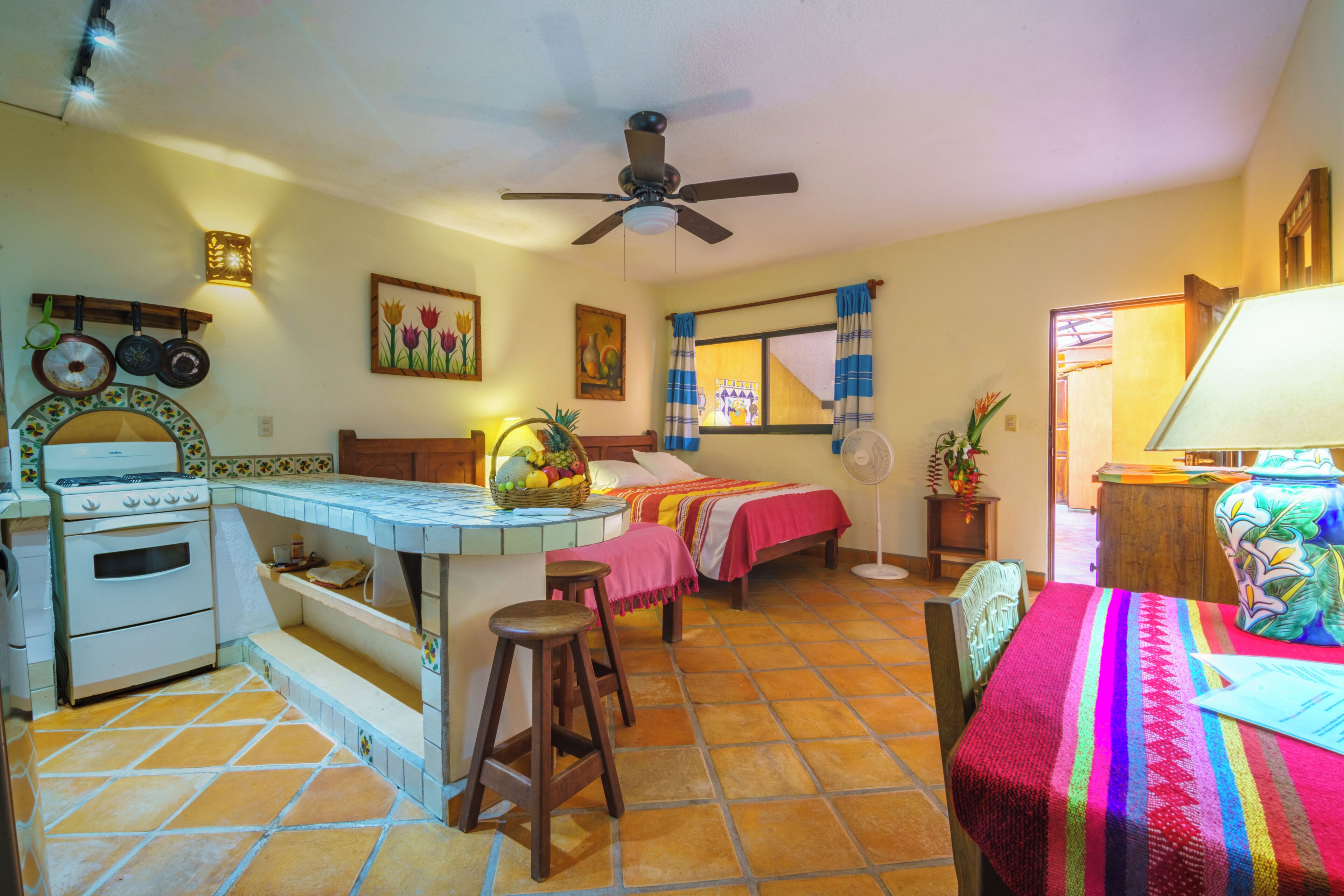 Cozinha compacta com fogão, área de refeições com banquetas, cama com manta colorida e ventilador de teto.