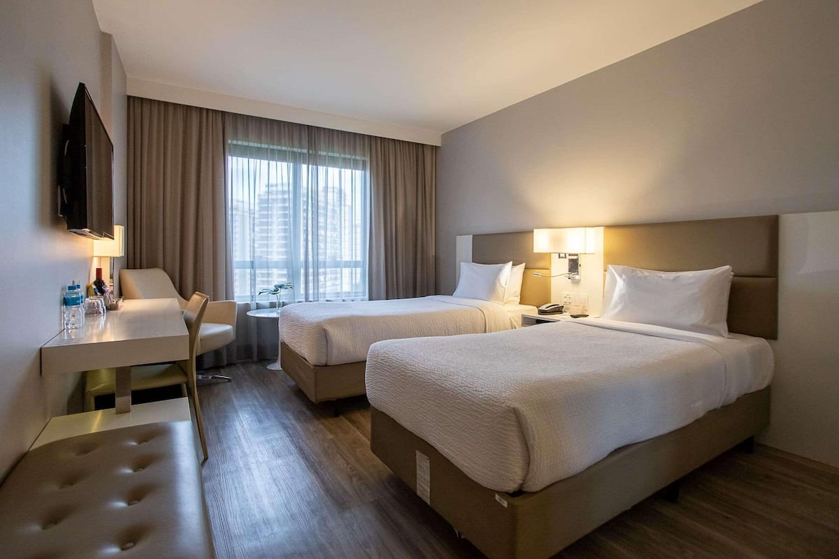 Quarto de hotel com duas camas, uma escrivaninha, uma cadeira e uma televisão.