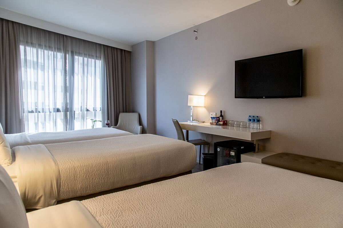 Quarto de hotel com duas camas, uma mesa com luminária, televisão e janela com cortinas.