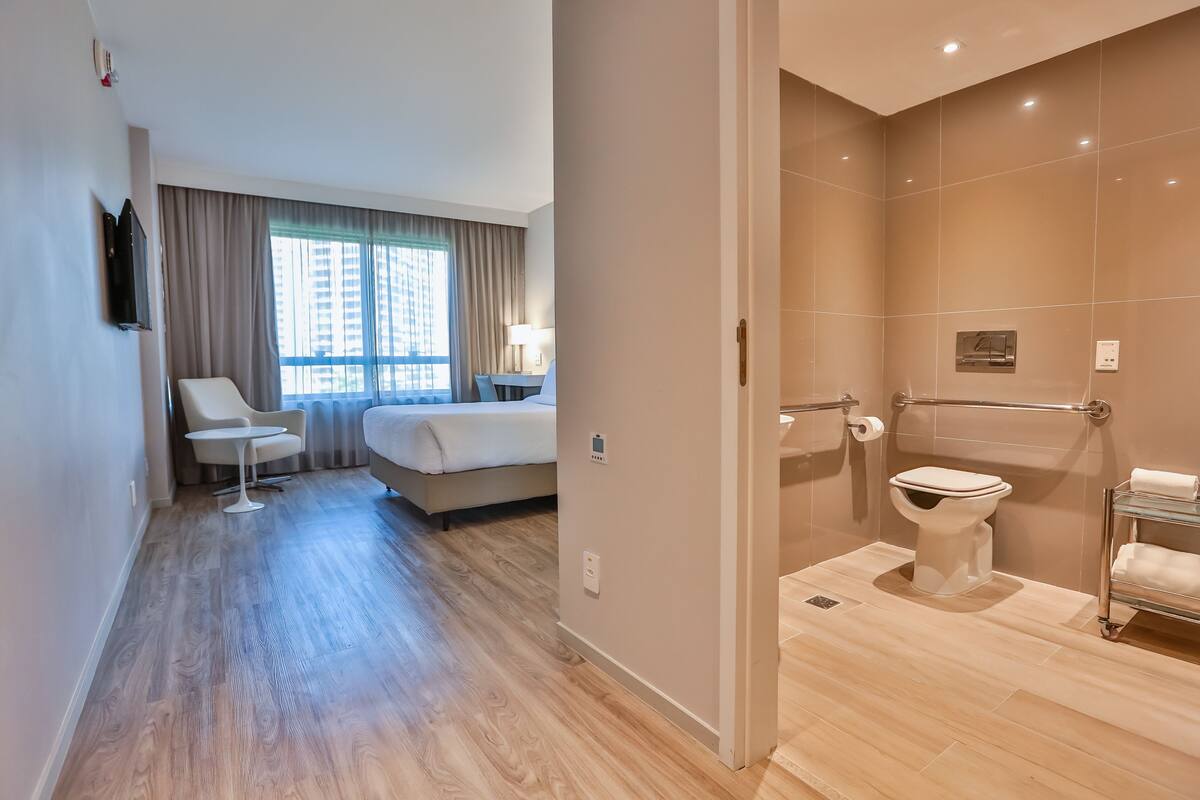 Quarto de hotel com uma cama, uma mesinha, uma cadeira, uma televisão e um banheiro com vaso sanitário e box de vidro.