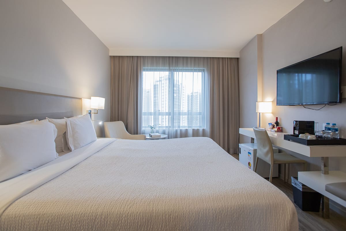 Quarto de hotel com uma cama grande, uma mesa com computador, televisão e janela com cortinas.