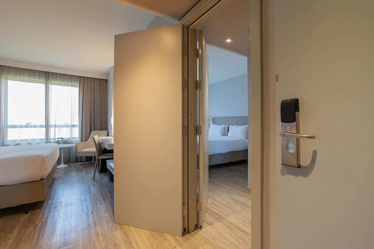 Quarto de hotel com cama, escrivaninha e cadeira. Há uma porta com fechadura e leitor de cartão.