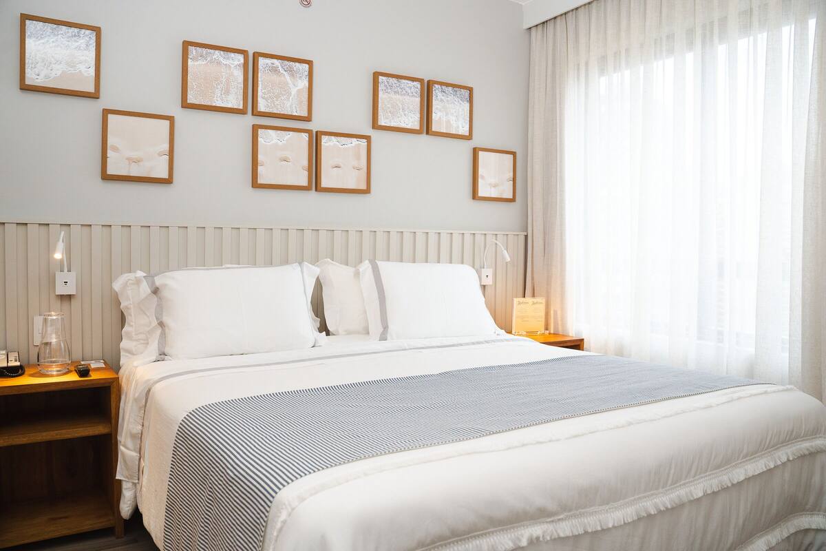 Quarto de hotel bem arrumado, com uma cama grande, mesa de cabeceira com abajur e parede decorada com quadros.