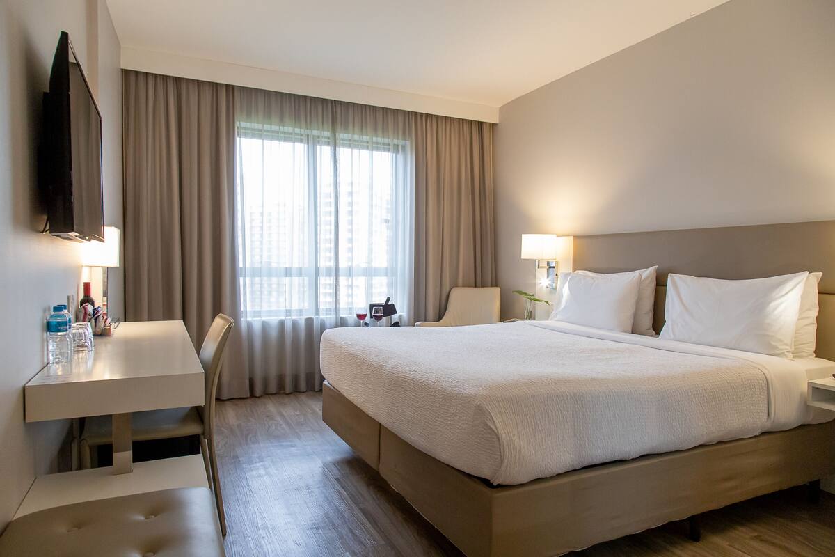Quarto de hotel com uma cama grande, uma escrivaninha com cadeira, uma TV na parede e uma janela com cortinas.