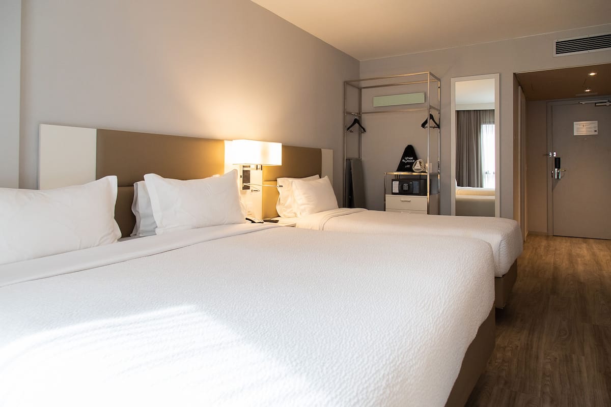 Quarto de hotel com duas camas, uma mesa de cabeceira com abajur, um espelho, um guarda-roupa e uma porta.