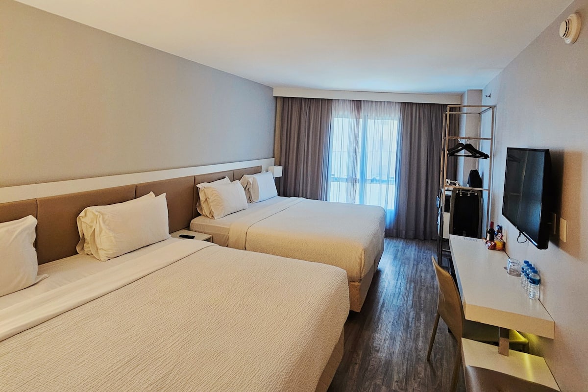 Quarto de hotel com duas camas, uma escrivaninha, uma televisão e um guarda-roupa.