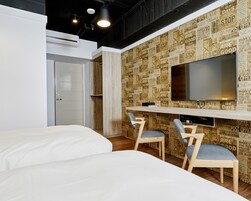 Quarto de hotel moderno com uma cama, uma mesa de escritório com duas cadeiras, uma televisão grande e uma parede com impressão de jornais antigos.